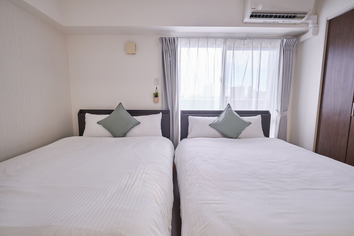 Quarto duplo com duas camas, janela com cortinas, ar condicionado fixado na parede e uma pequena mesa de cabeceira com abajur.