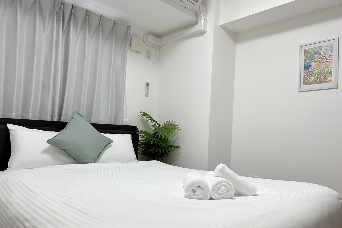 Cama bem arrumada com lençóis brancos, um travesseiro verde e toalhas dobradas.