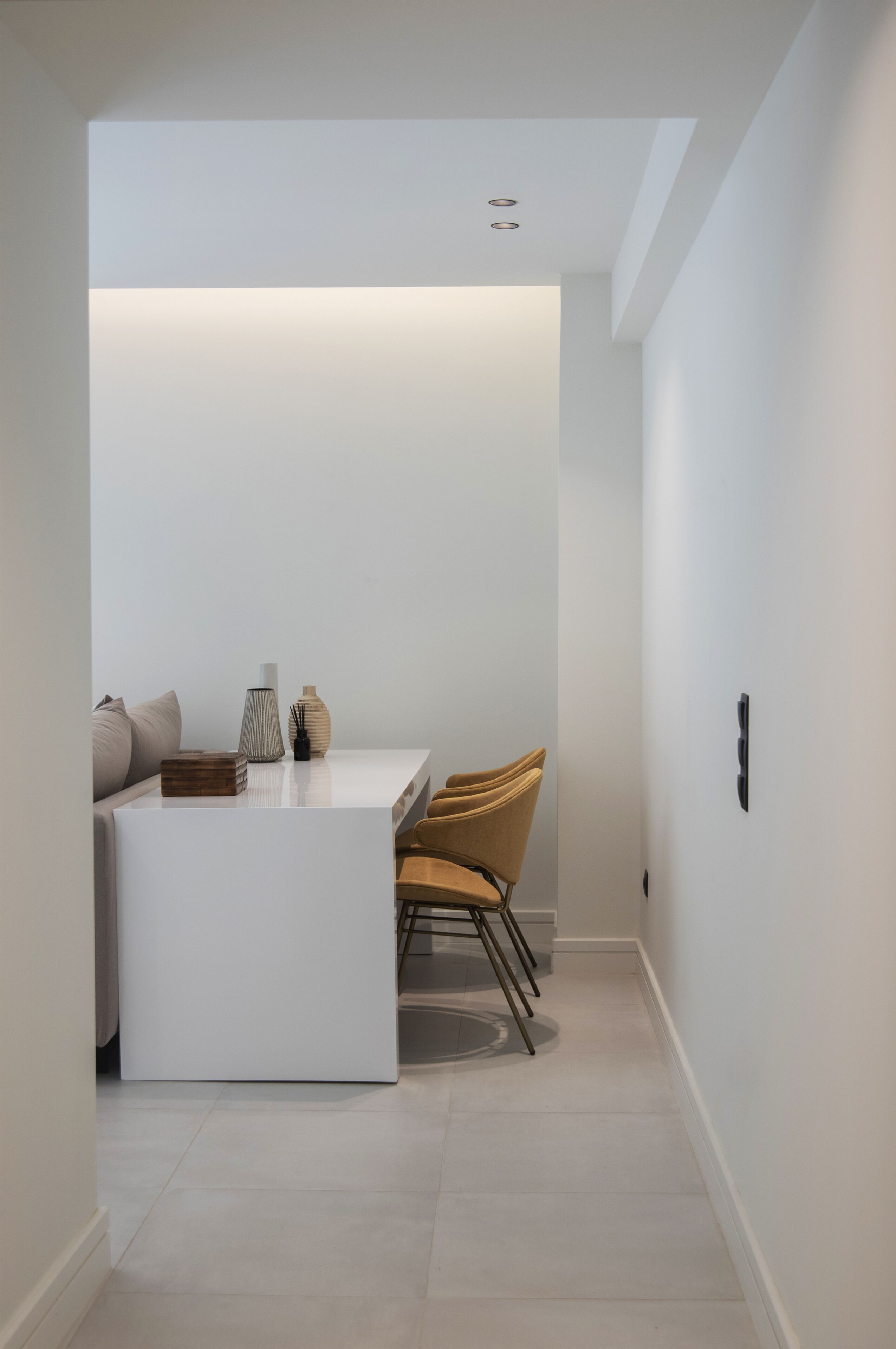 Um corredor estreito com um sofá branco, uma mesinha com objetos decorativos e uma cadeira.