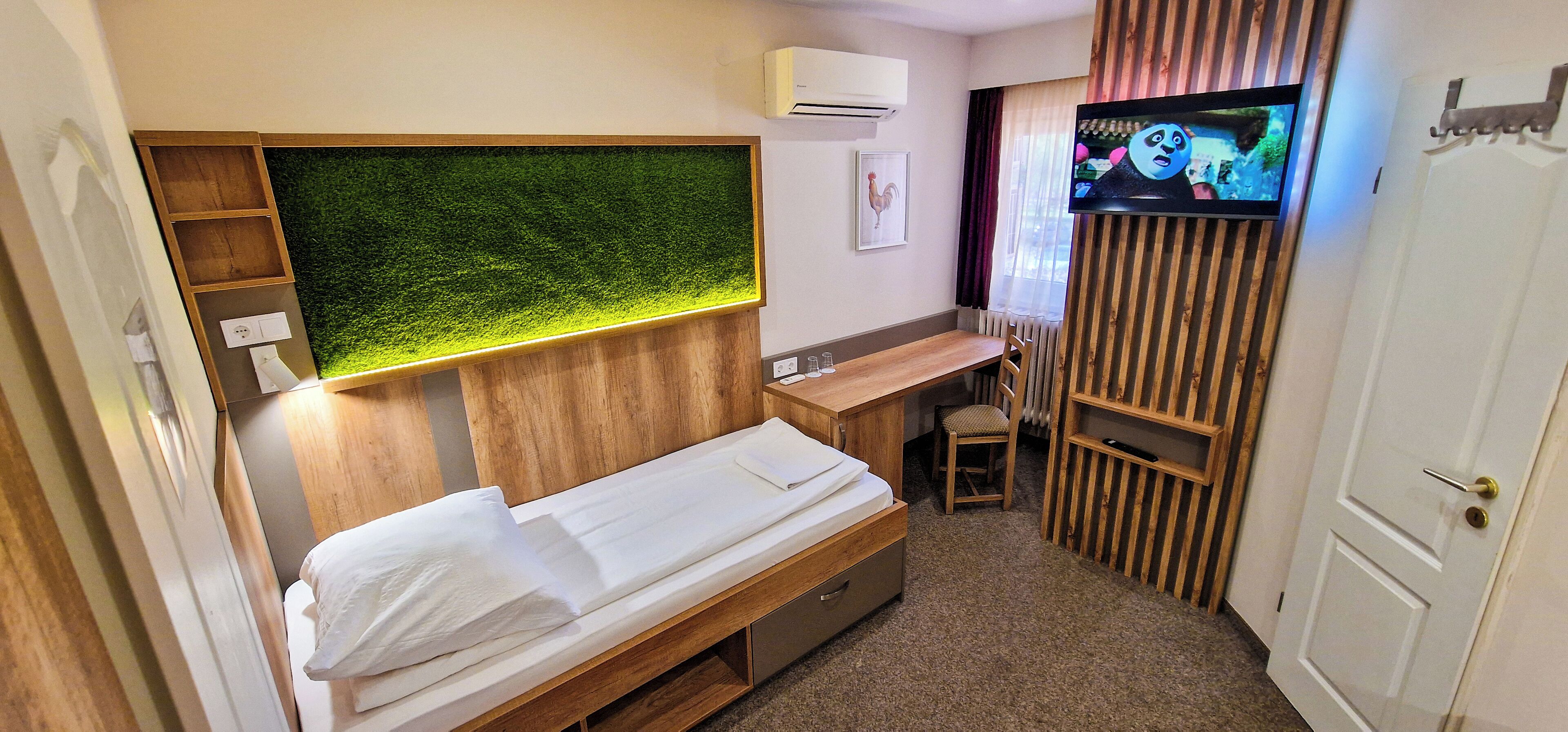 Quarto de hotel compacto com uma cama, uma secretária pequena, uma televisão e ar condicionado fixado na parede.