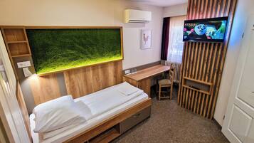 Quarto de hotel compacto com uma cama, uma secretária pequena, uma televisão e ar condicionado fixado na parede.