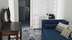 Um espaço de estar compacto com um sofá azul, uma mesinha de madeira com um espelho, um guarda-roupa branco e um chão revestido de azulejos com padrões geométricos.