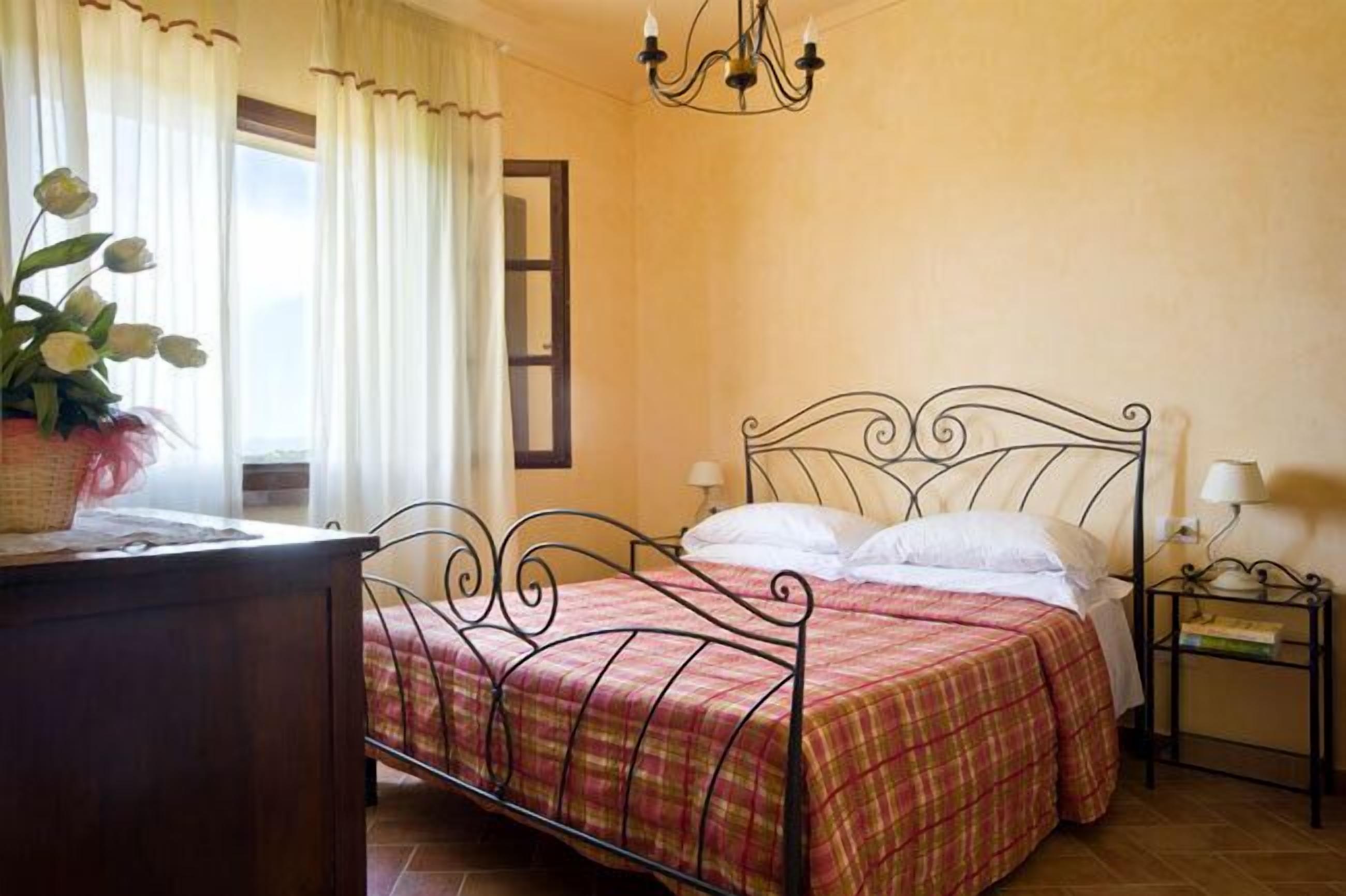 Quarto com cama, cortinas, lustre, mesa de cabeceira e abajur.