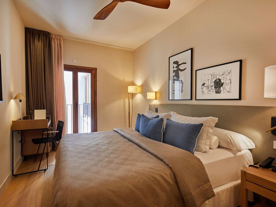 Quarto de hotel com cama, escrivaninha, cadeira, abajur, obras de arte e ventilador de teto.