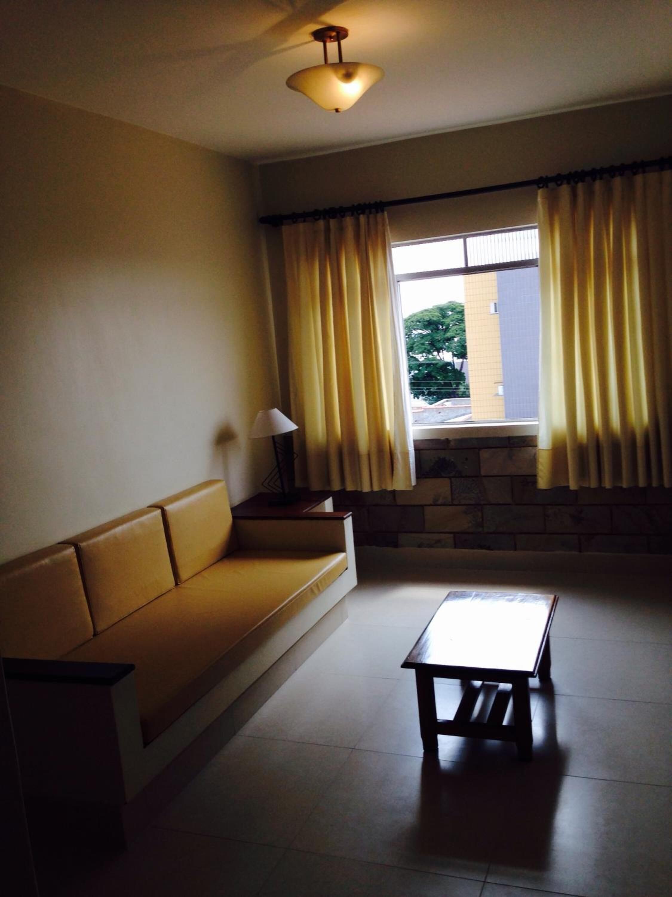 Sala de estar com sofá, mesa de centro e luminária. Há uma janela com cortinas e vista para edifícios lá fora.