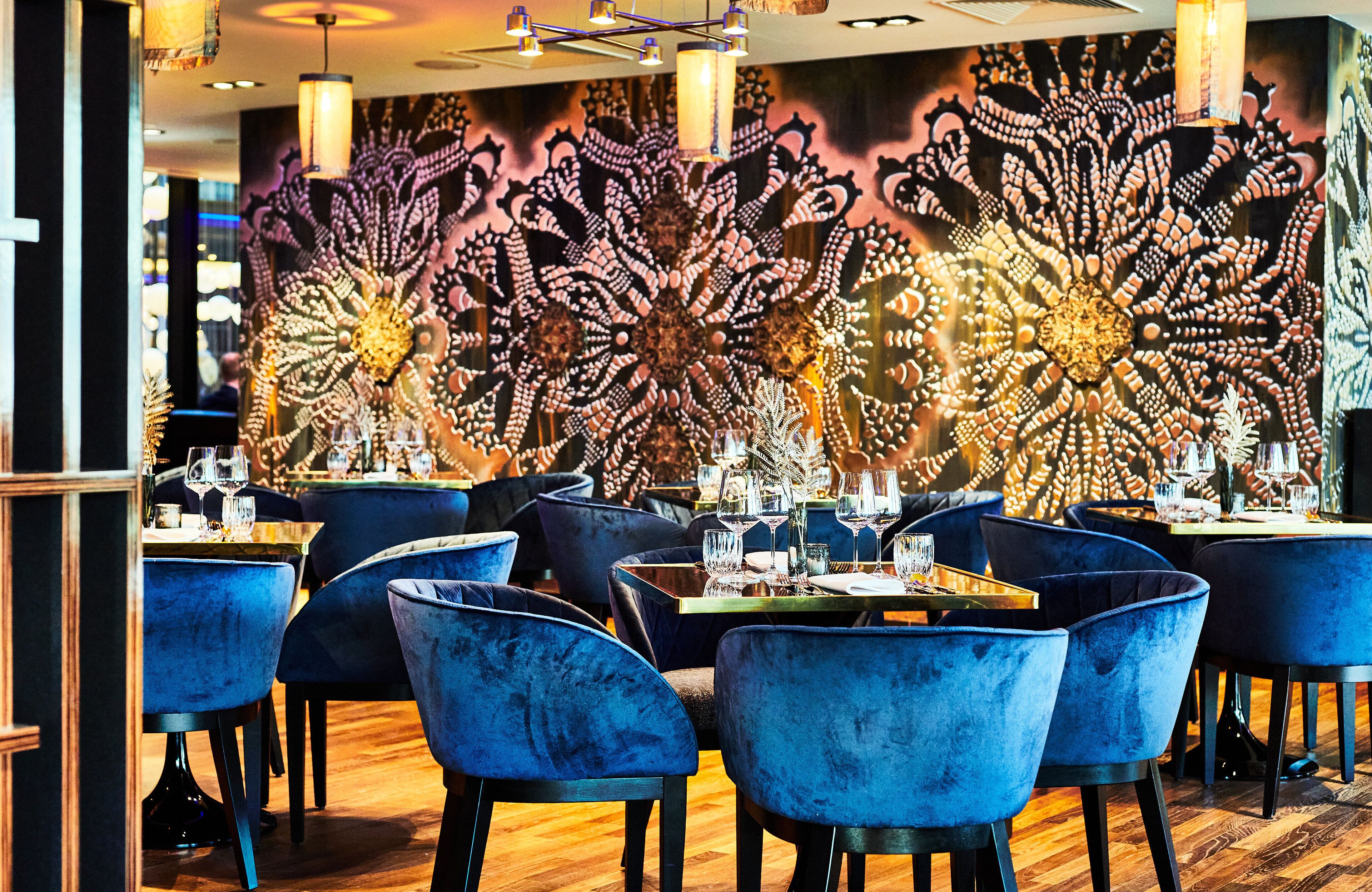 Interior de restaurante com cadeiras de veludo azul, mesas postas com copos de vinho e uma grande parede decorativa com padrões elaborados.