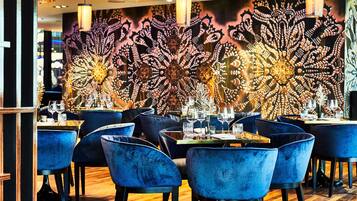 Interior de restaurante com cadeiras de veludo azul, mesas postas com copos de vinho e uma grande parede decorativa com padrões elaborados.