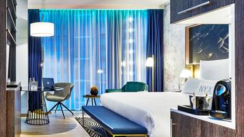 Quarto de hotel moderno com uma cama grande, uma escrivaninha com um laptop, uma cadeira, um banco e vista para a cidade através de cortinas leves.