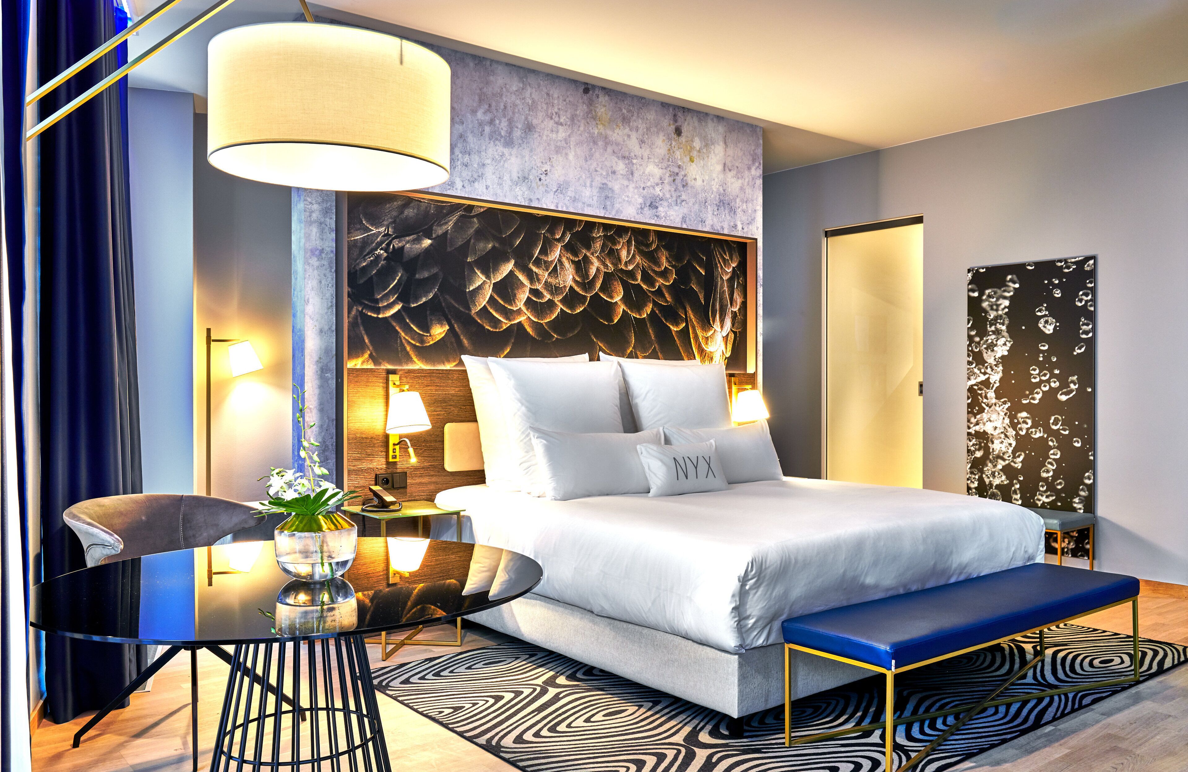 Quarto de hotel moderno com uma cama grande, uma mesa de cabeceira com abajur, uma cadeira, uma pequena mesa com um vaso de flores e um grande quadro abstrato na parede.