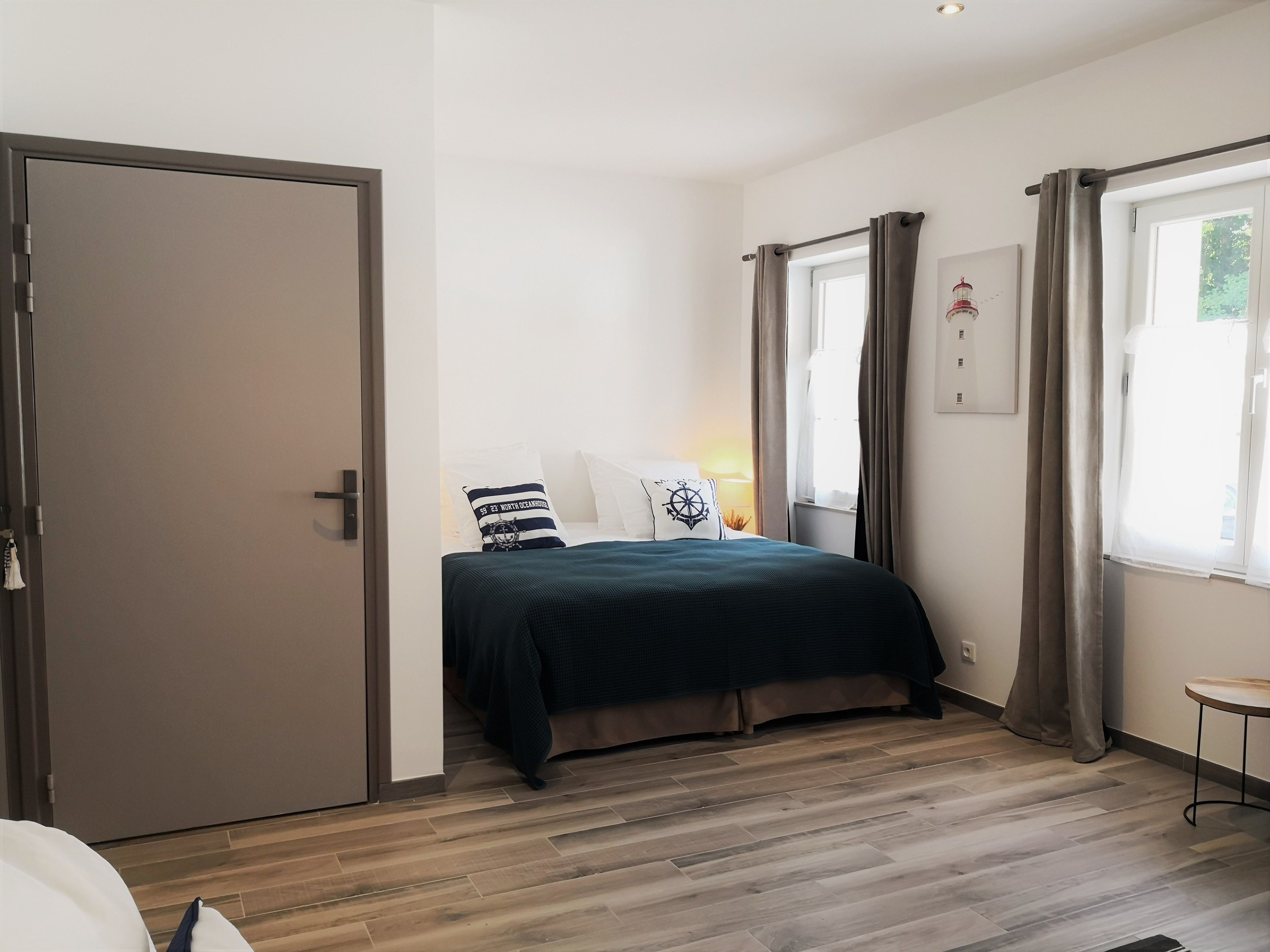 Quarto com cama, porta, janela com cortinas e uma pequena mesa.