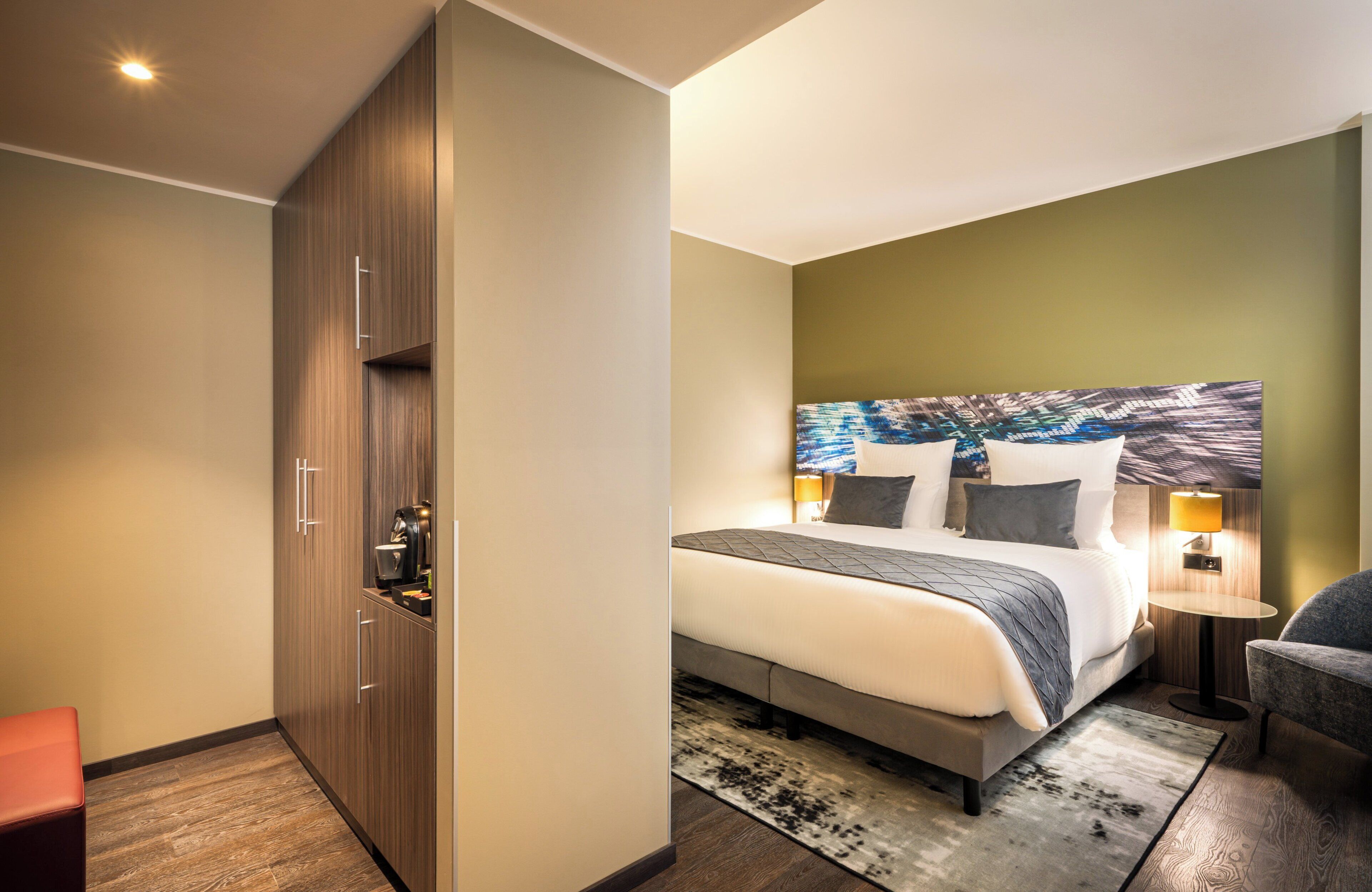 Quarto de hotel moderno com uma cama grande, uma mesa de cabeceira com abajur, um espaço de estar com uma poltrona e um guarda-roupa.