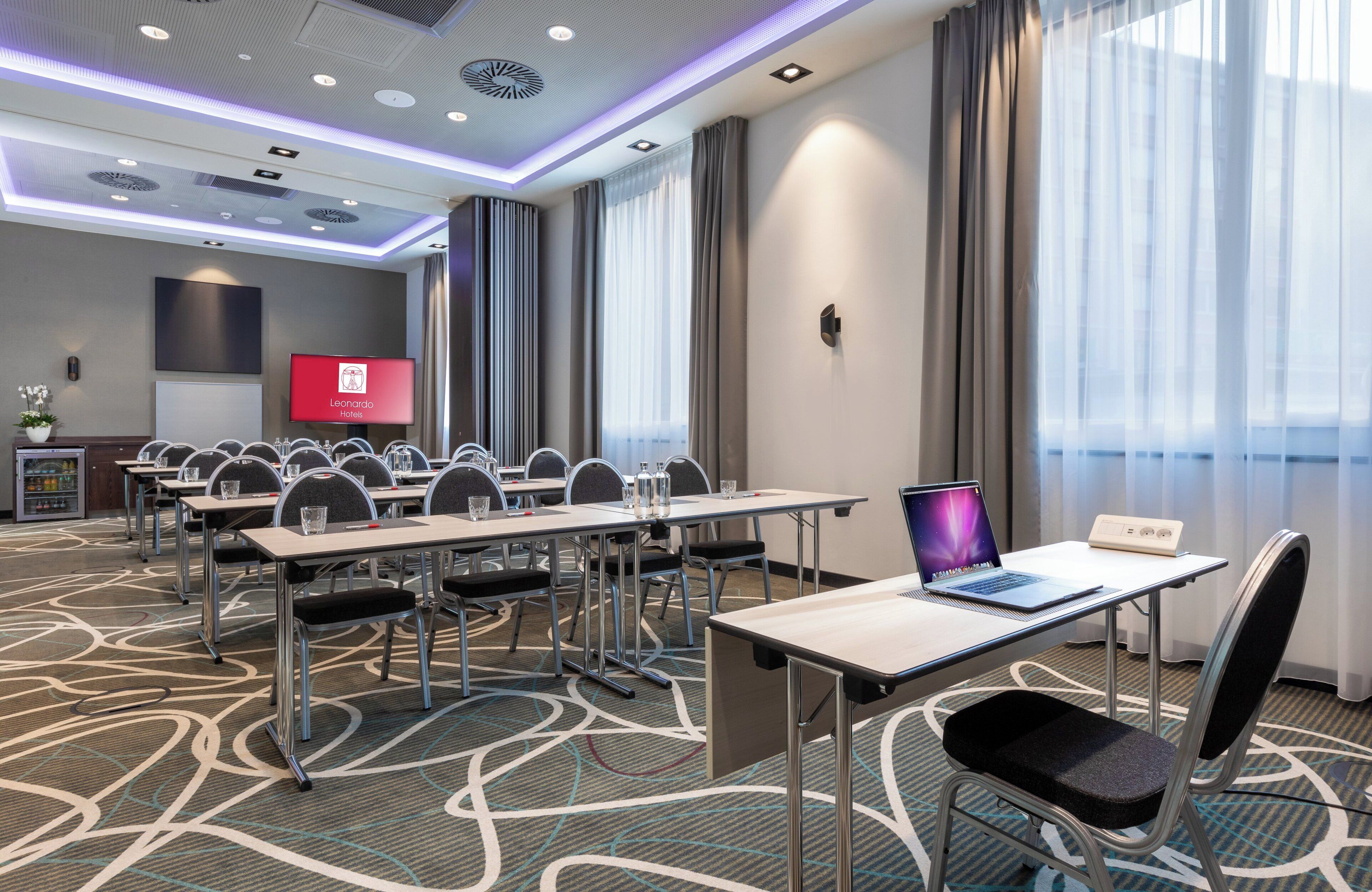 Uma sala de conferências de hotel com várias mesas, cadeiras, uma tela de televisão e uma janela grande com cortinas.