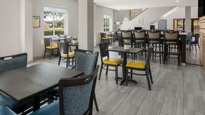 Uma área de jantar moderna com várias mesas e cadeiras, uma TV de tela plana na parede e vista para o exterior através de grandes janelas.