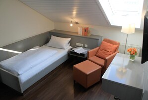 Quarto de hotel pequeno com duas camas de solteiro, uma mesinha, uma cadeira e uma mesinha de cabeceira com livros.