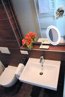 Um banheiro com pia branca, vaso sanitário, bidê, espelho iluminado e um vaso com flores.