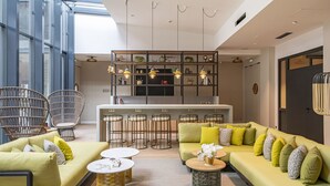 Uma sala de estar moderna com sofá, cadeiras e mesa de centro. Há uma área de cozinha com balcão e prateleiras com plantas.