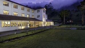 Um edifício grande e branco com várias janelas e uma galeria coberta, rodeado por um jardim bem cuidado e árvores.