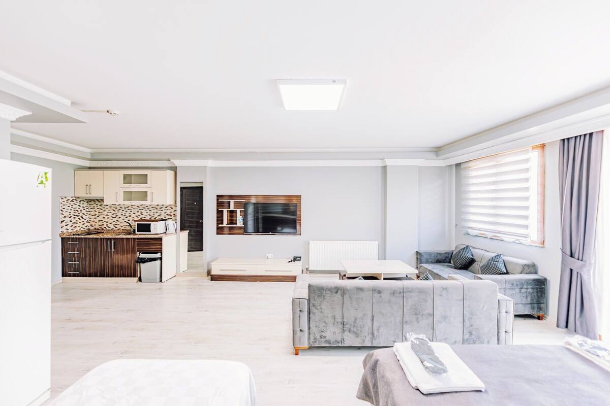 Uma sala de estar moderna com um sofá cinzento, uma mesinha e uma televisão fixada na parede. Há uma área de cozinha com armários e um micro-ondas.