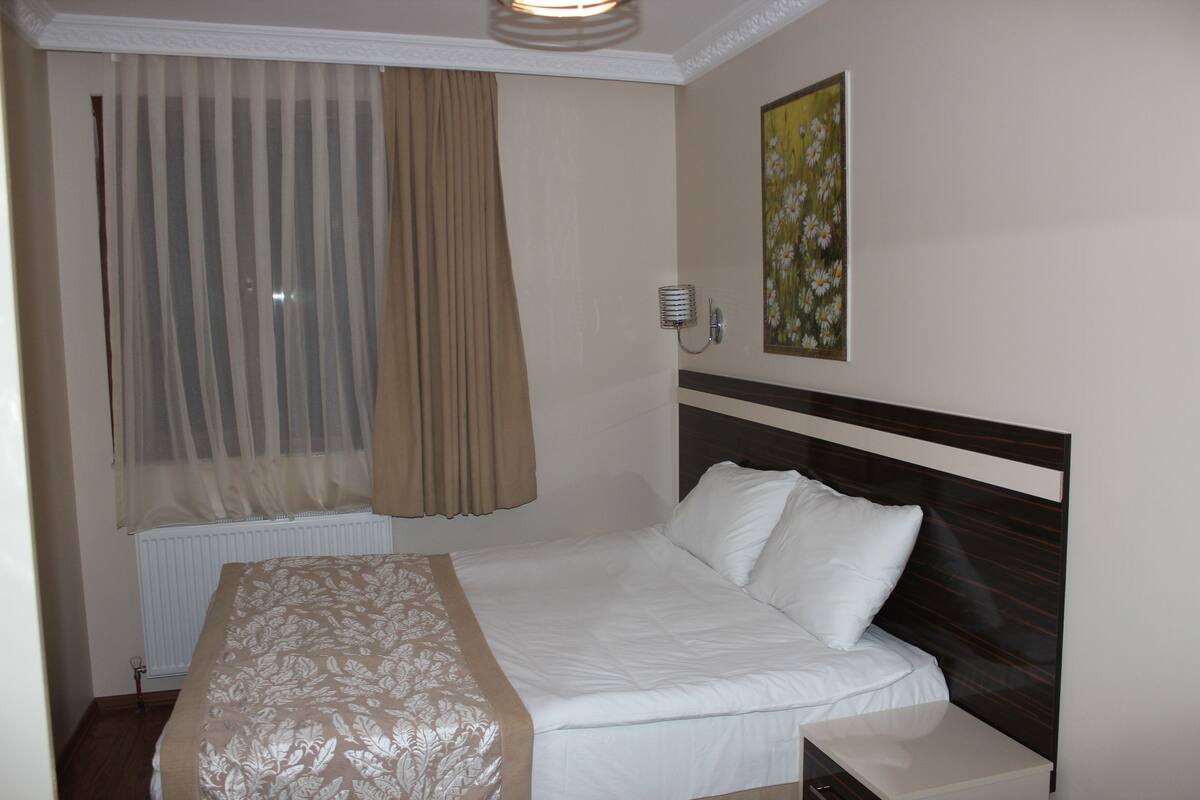 Quarto de hotel com cama, mesa de cabeceira, janela com cortinas, radiador e luminária fixada na parede.
