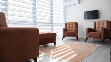 Uma sala de estar com uma janela grande, uma TV de tela plana e três poltronas marrons.
