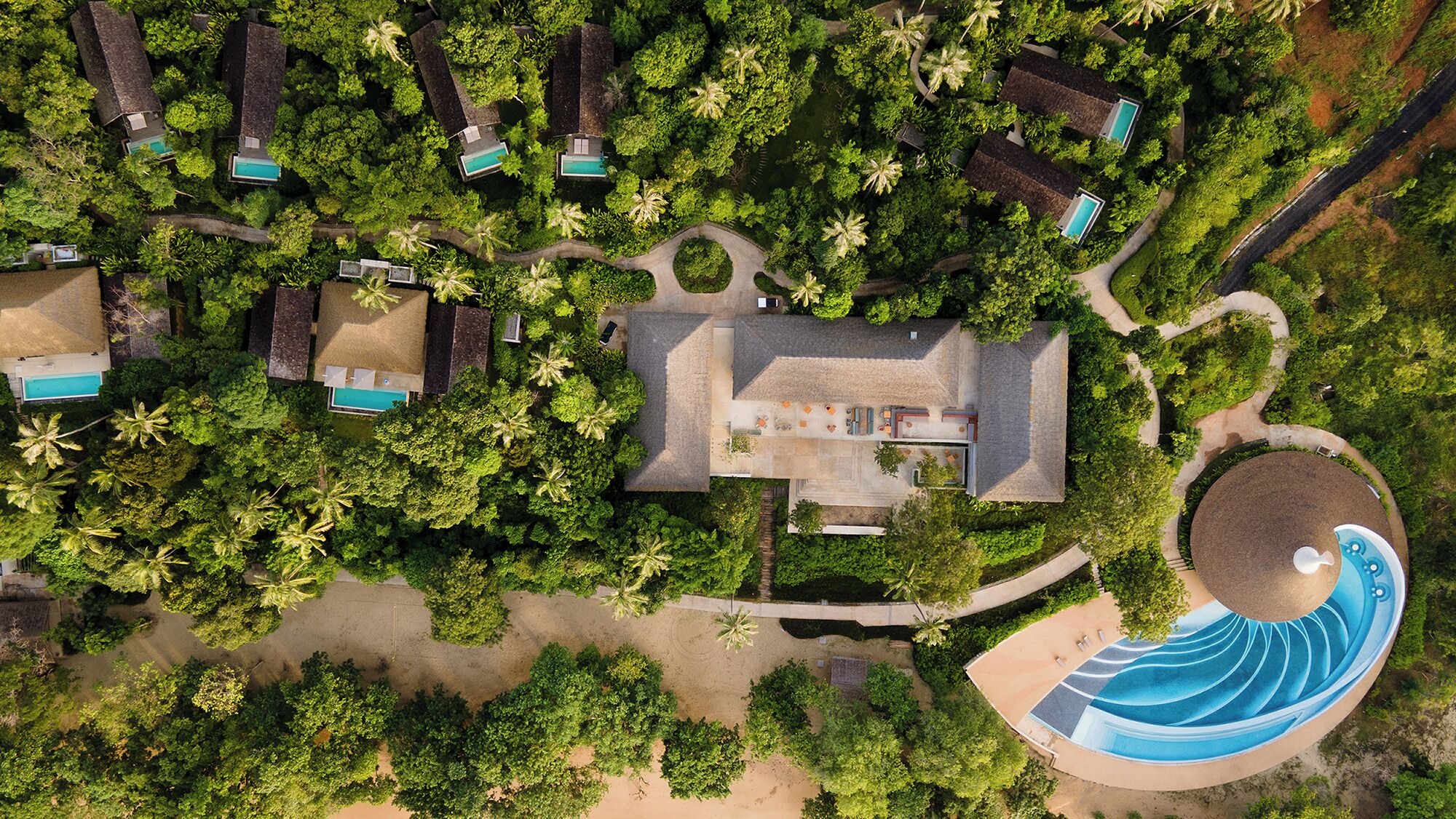 Vista aérea de um resort com várias acomodações em estilo bangalô, um edifício central com piscina e uma grande piscina circular.