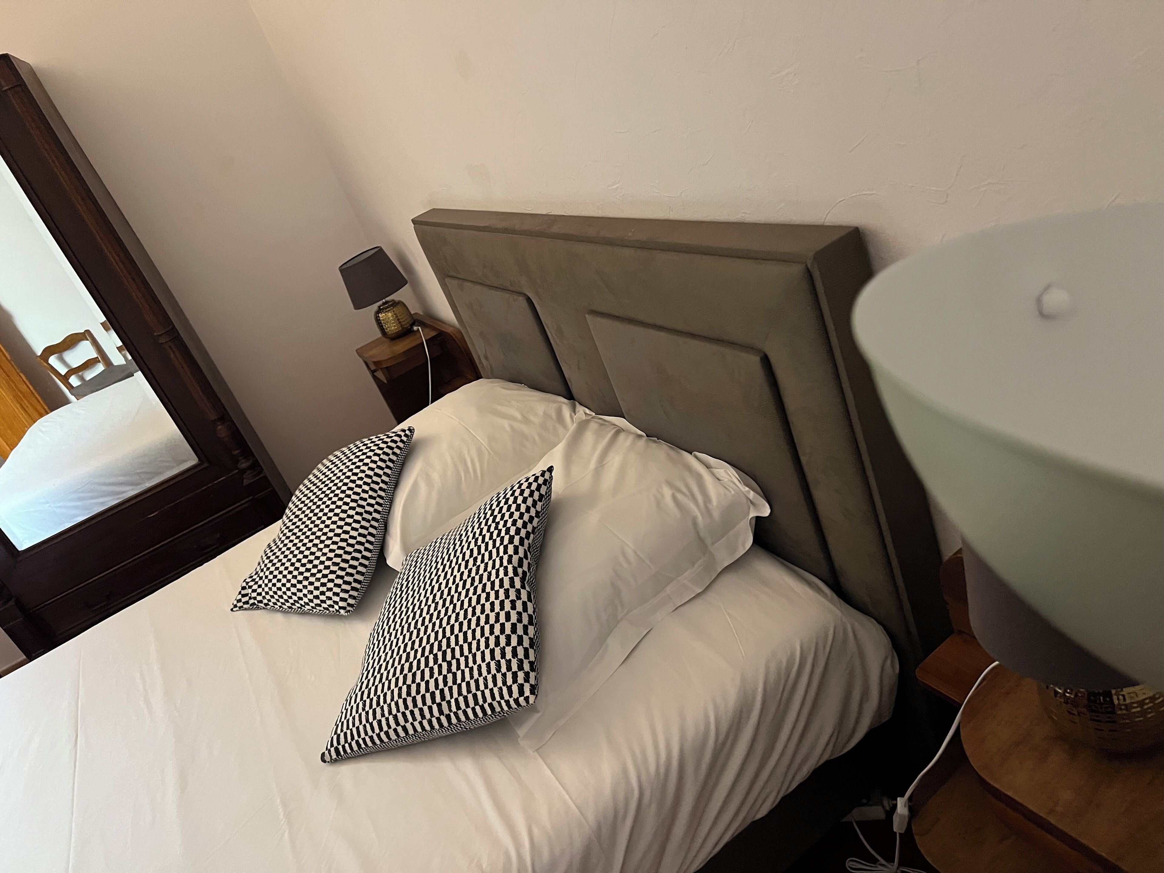 Cama bem arrumada com duas almofadas xadrez, luminária na mesa de cabeceira e uma mesa redonda com abajur.