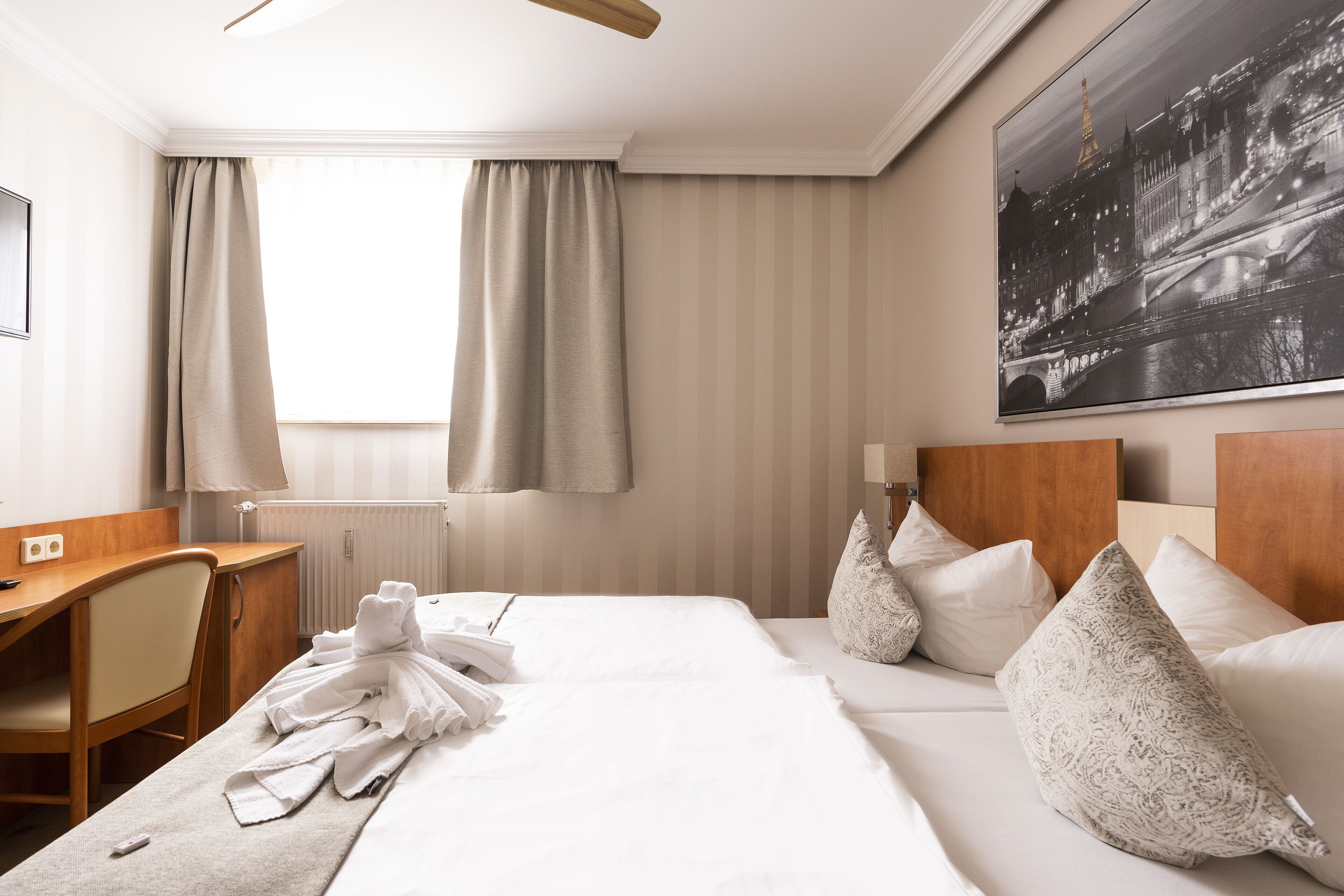 Quarto de hotel com uma cama, uma escrivaninha e uma cadeira. Há uma janela com cortinas, um radiador e um quadro na parede.