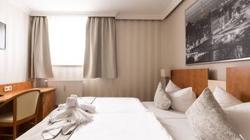 Quarto de hotel com uma cama, uma escrivaninha e uma cadeira. Há uma janela com cortinas, um radiador e um quadro na parede.