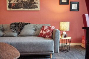 Uma sala de estar com um sofá cinzento, uma almofada floral vermelha, um mantê de pele, um abajur sobre uma mesa de arame e quadros nas paredes.