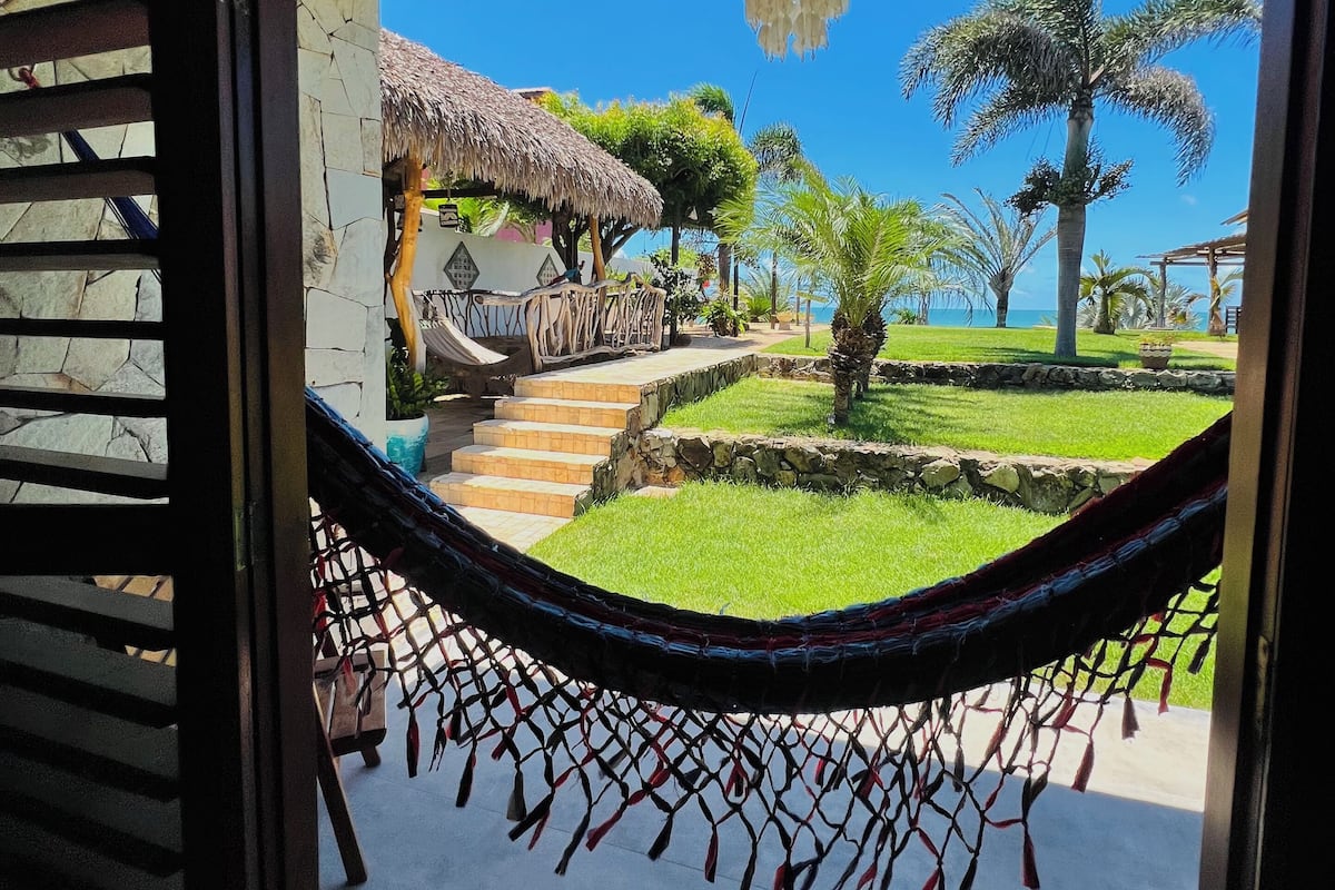 Uma rede em um quarto com vista para um jardim e uma construção com telhado de palha.