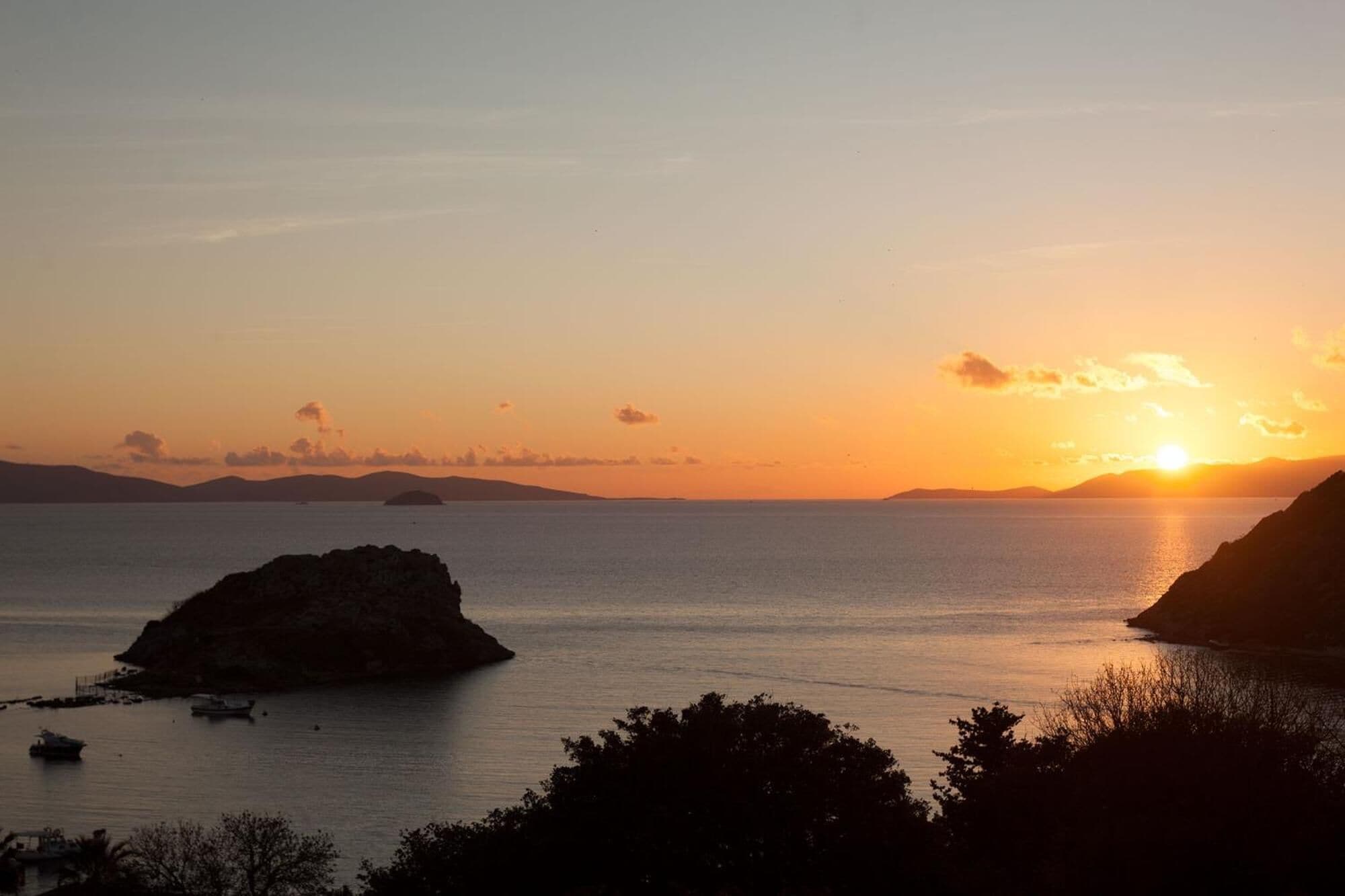 Vista de um pôr do sol costeiro com águas calmas, uma pequena ilha e montanhas ao longe.