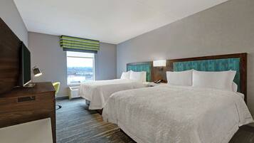 Quarto de hotel com duas camas, uma secretária com computador, janela com vista e cortina verde riscada.