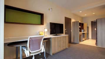 Quarto de hotel moderno com escrivaninha, cadeira, televisão e cozinha compacta.