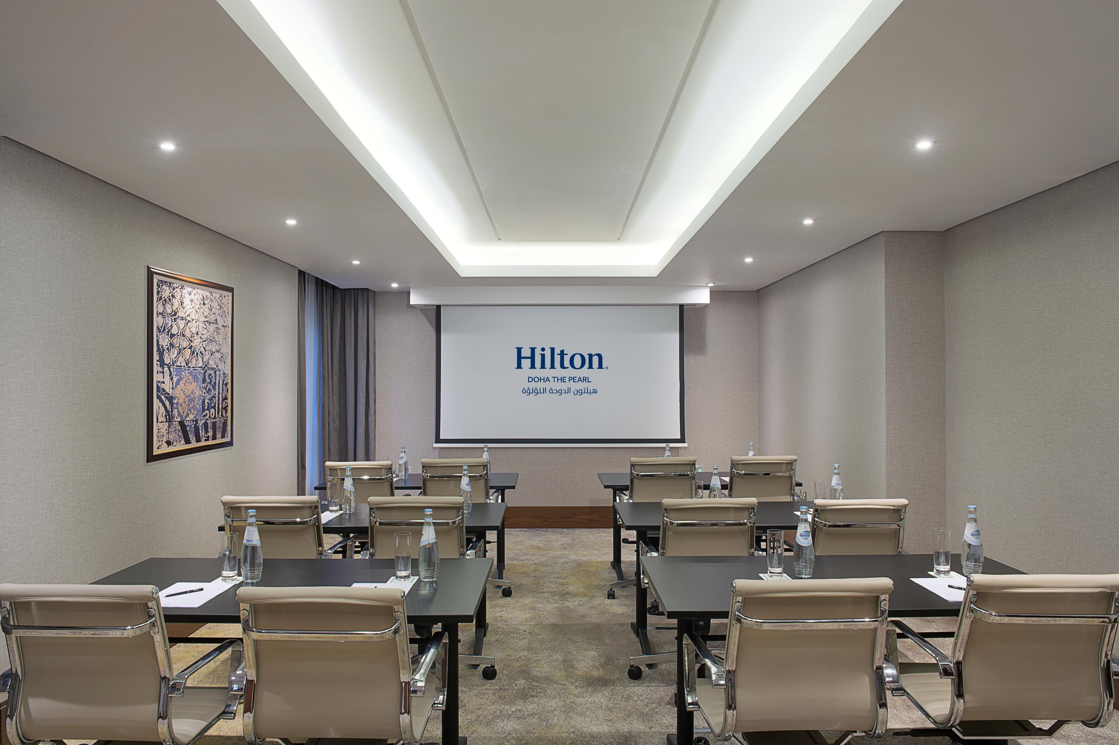 Uma sala de conferências com uma tela grande exibindo 'Hilton DOHA THE PEARL' em inglês e árabe, cadeiras bege, mesas com garrafas de água e piso carpeteado.