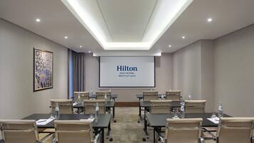 Uma sala de conferências com uma tela grande exibindo 'Hilton DOHA THE PEARL' em inglês e árabe, cadeiras bege, mesas com garrafas de água e piso carpeteado.