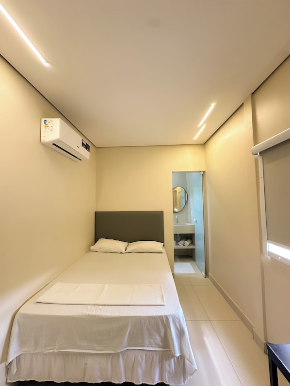 Quarto de hotel pequeno e moderno, com uma cama de solteiro, mesa de cabeceira, espelho e ar condicionado integrado.