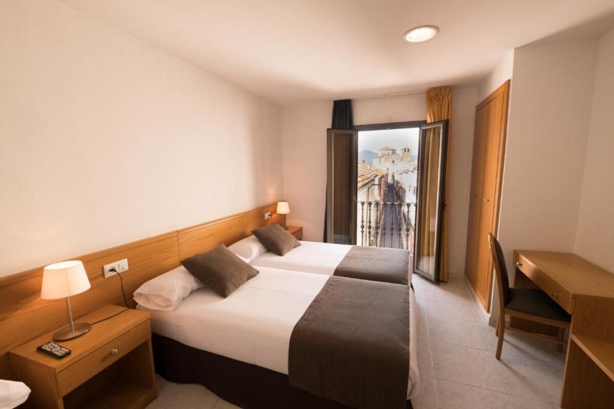 Quarto de hotel com duas camas, uma mesa de trabalho e uma varanda com vista para a cidade.