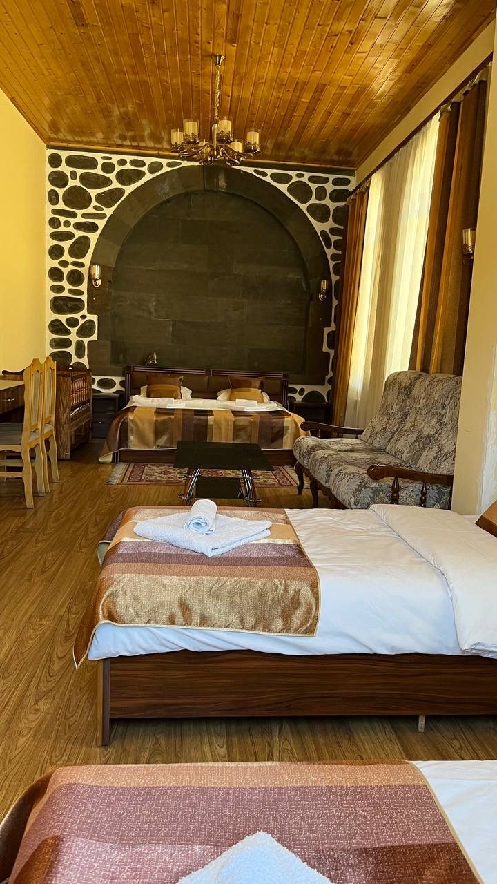 Quarto de hotel com lareira de pedra, teto de madeira, cama com colcha estampada, um sofá e uma cadeira.