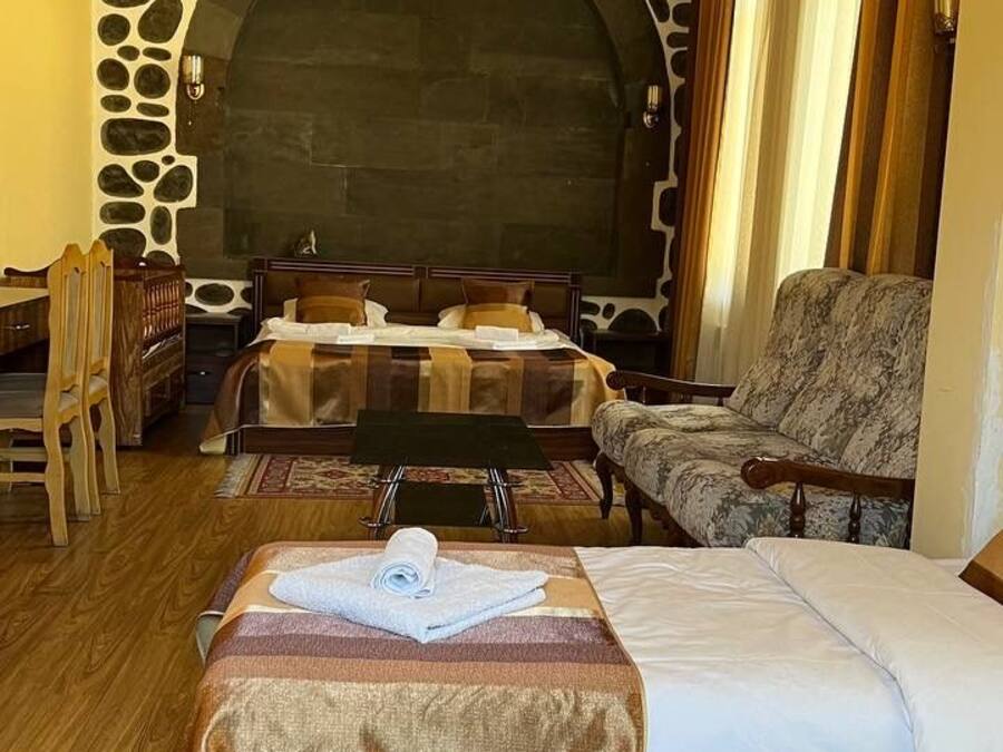 Quarto de hotel com lareira de pedra, teto de madeira, cama com colcha estampada, um sofá e uma cadeira.