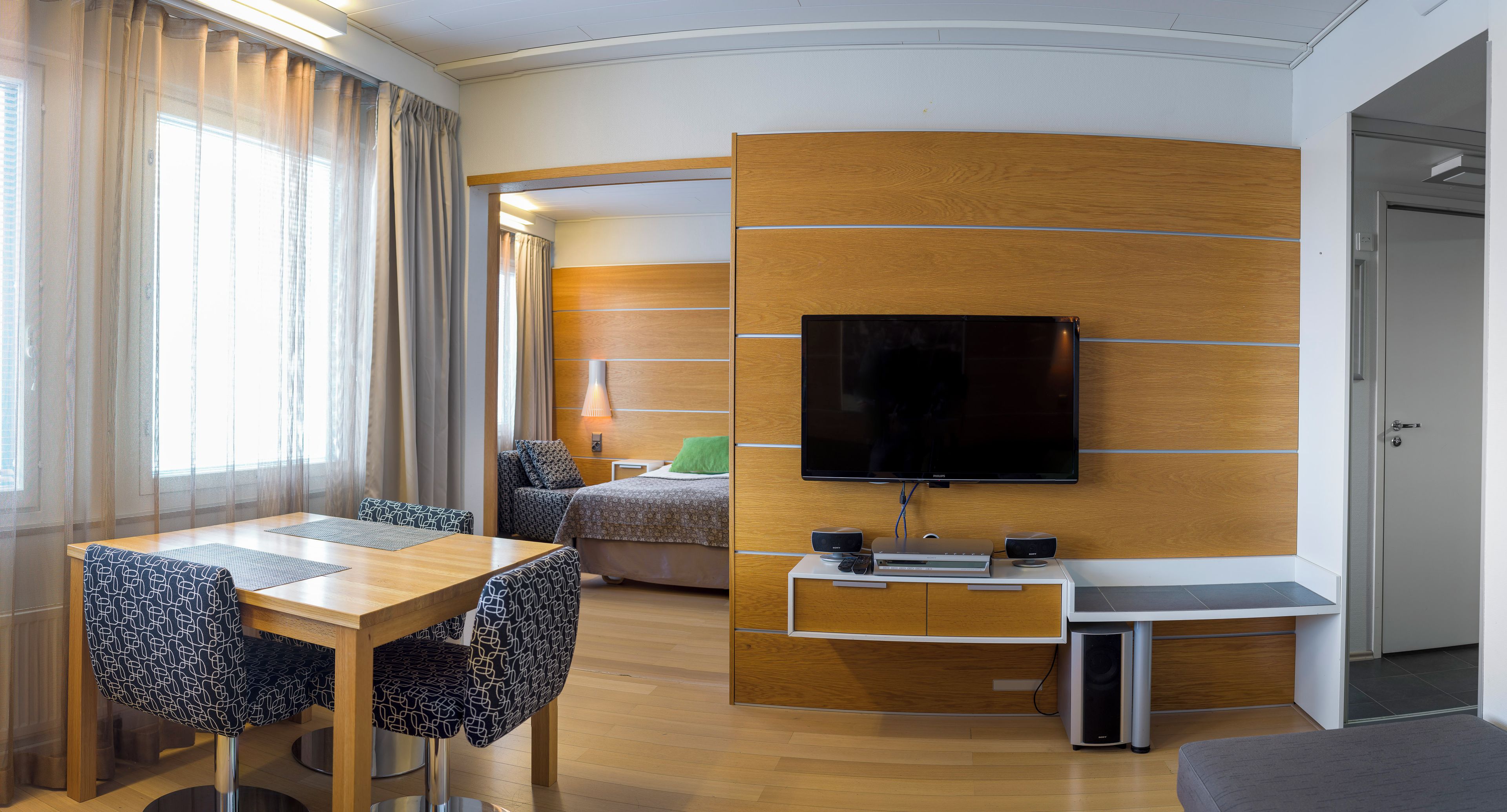 Quarto de hotel com área de refeições, televisão fixada em um painel de madeira e uma cama ao fundo.