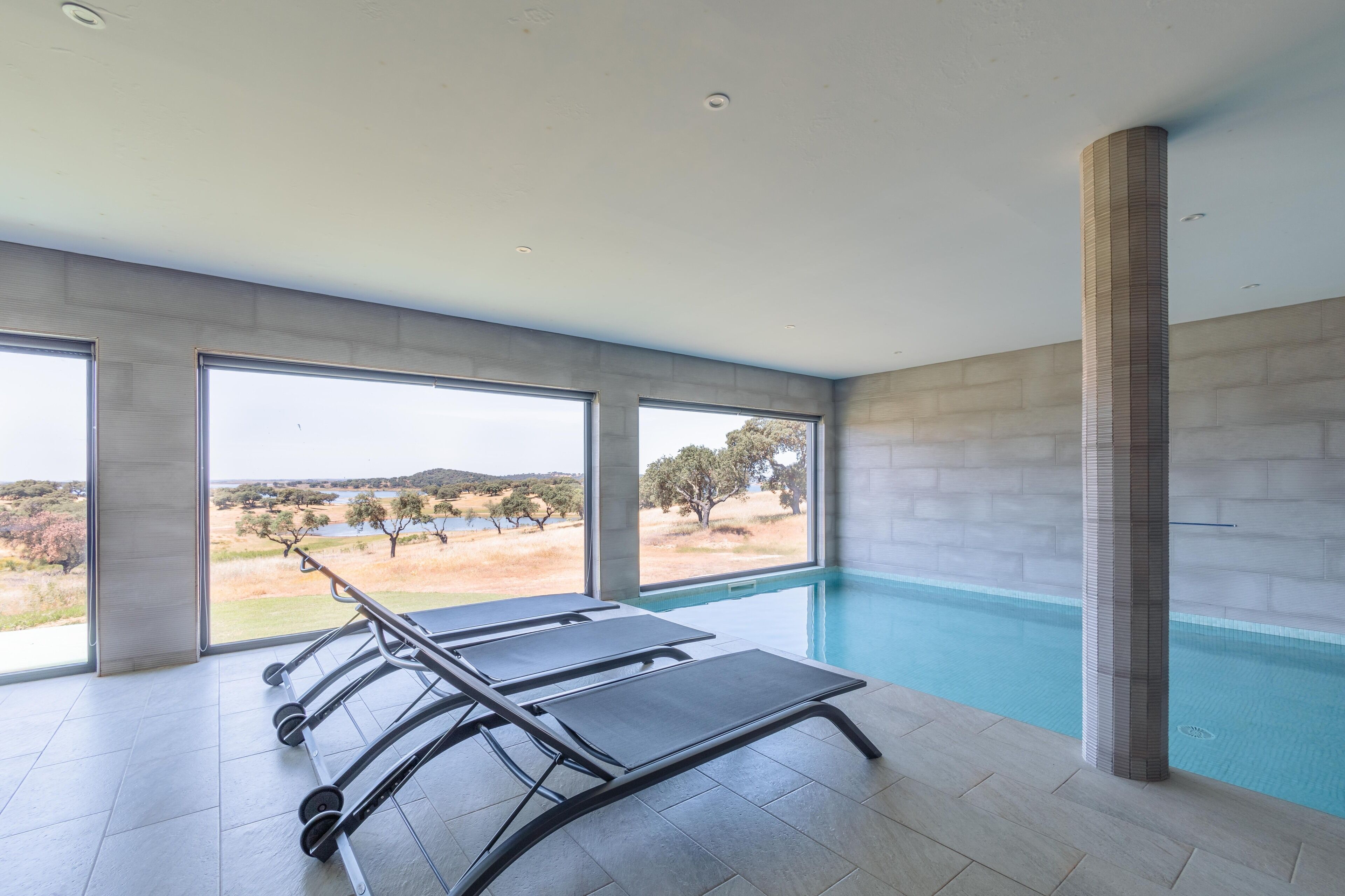 Uma área moderna de piscina interna com amplas janelas, vista para o exterior e um espaço de descanso à beira da piscina.