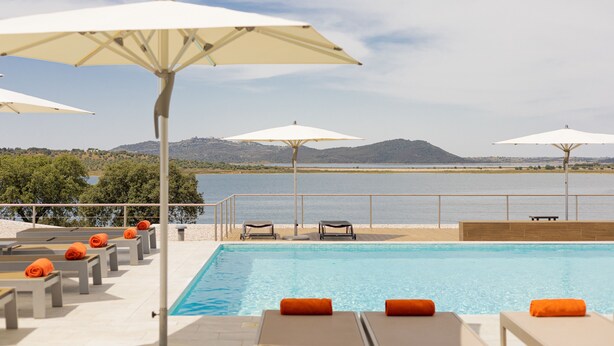 Área de piscina com espreguiçadeiras e guarda-sóis, com vista para um corpo de água e colinas.