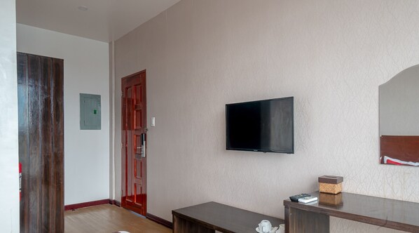 Quarto com televisão de tela plana na parede, porta de madeira, mesa pequena com xícara e pires, e um banco.