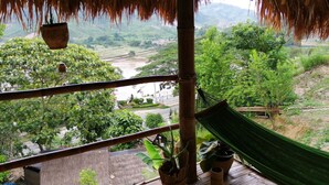 Uma rede sob um telhado de palha, com vista para um vale exuberante, um rio e montanhas ao longe.