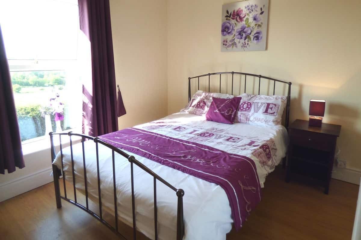 Quarto com cama, mesa de cabeceira, janela com cortinas e um quadro floral na parede.
