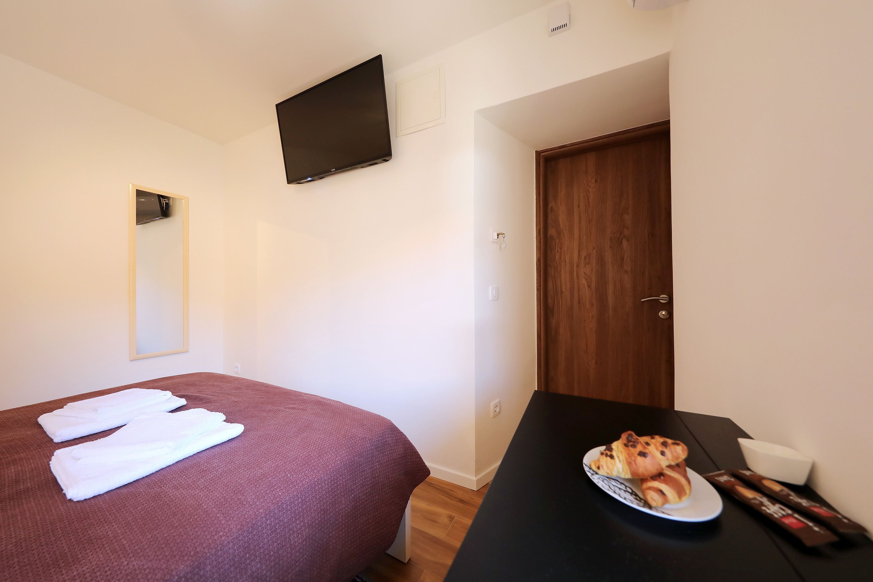 Quarto de hotel com uma cama, televisão de tela plana na parede, porta de madeira e uma mesa com croissants e uma xícara de café.