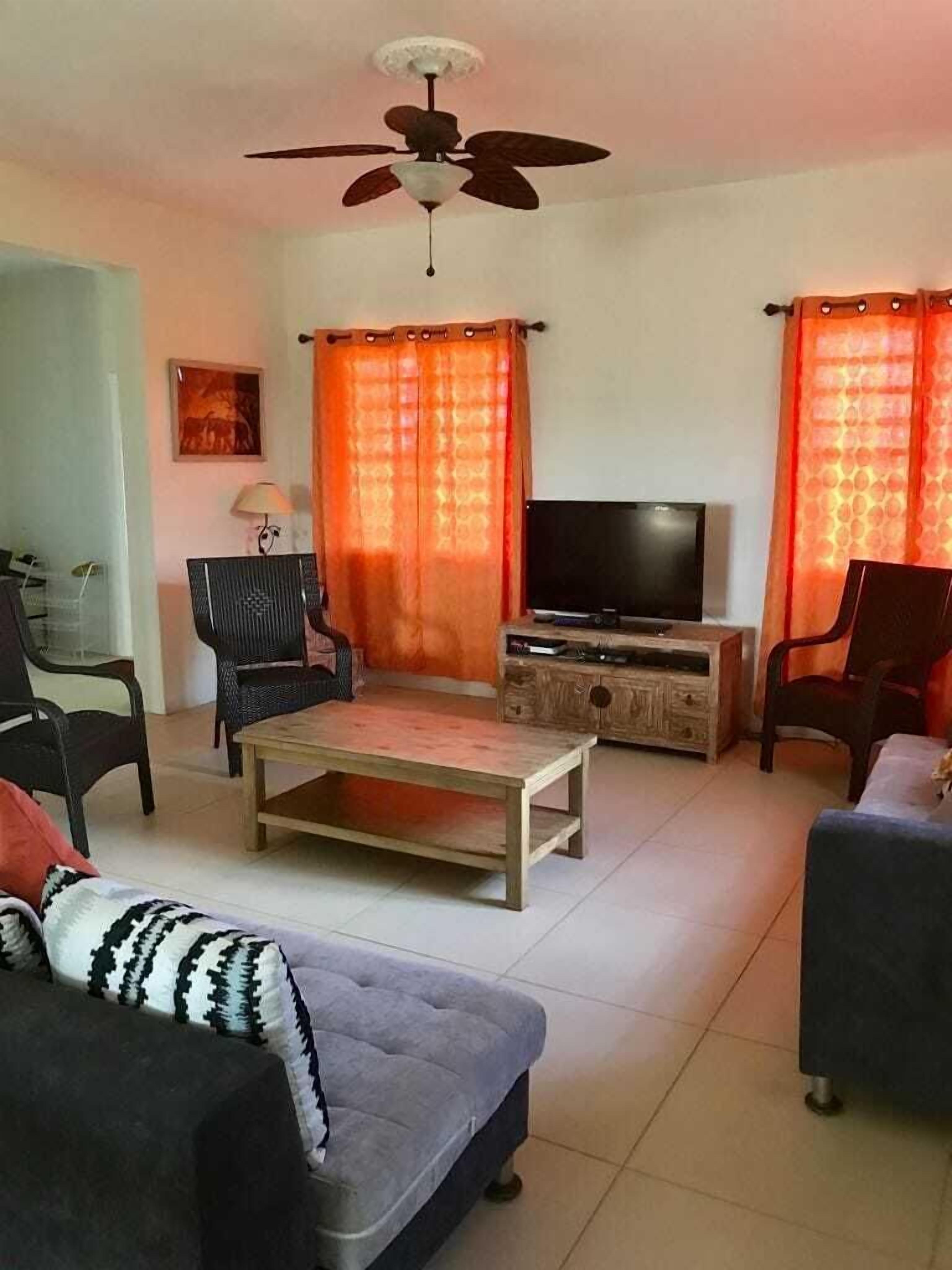 Sala de estar com sofá, cadeiras, mesa de centro, televisão e cortinas.