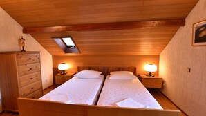 Quarto com duas camas de solteiro, cabeceiras de madeira, mesas de cabeceira com abajures, cômoda de madeira e teto inclinado com claraboia.