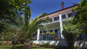 Um edifício de dois andares com varanda, cercado por vegetação exuberante e um gramado bem cuidado.