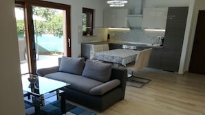Uma sala de estar moderna com um sofá cinzento, uma mesa de centro de vidro e uma pequena mesa de jantar com toalha de mesa estampada. O espaço possui piso de madeira clara, uma cozinha com armários brancos e vista para uma piscina através de uma porta de correr de vidro.